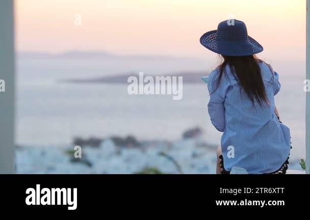 Young woman is looking at the sunset over a sea in famous village Mykonos with the old buildings ...