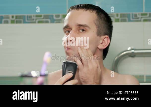 Young attractive man shaving with an electric razor in the bathroom. He ...