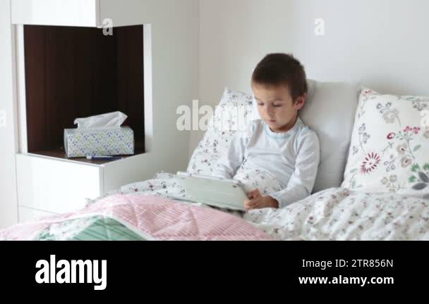 Sick child boy lying in bed with a fever,resting, playing on tablet ...