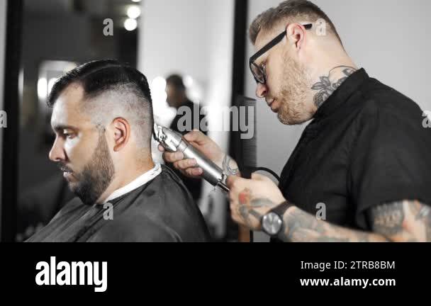 Tattoed barber makes haircut for customer at the barber shop by using ...