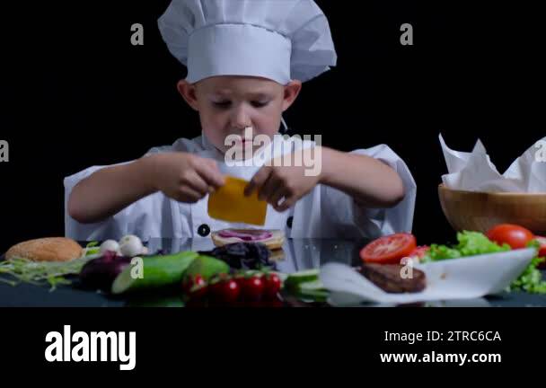 Cute boy is eating a burger while cooking. He wears chefs suit and cap ...