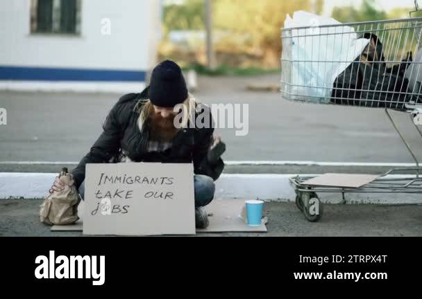 Angry upset young homeless man with cardboard sitting near shopping ...
