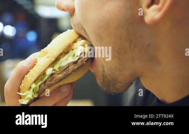 Profile of young man eating big hamburger or cheeseburger at food court ...
