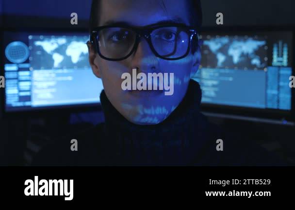 Portrait of Hacker programmer in glasses looks at camera while blue code characters reflect on ...