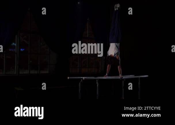 Male gymnast acrobat performs handstand on parallel bars in a dark room ...