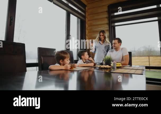 Family of four having Breakfast in his kitchen with large Windows. People are smiling, mother ...