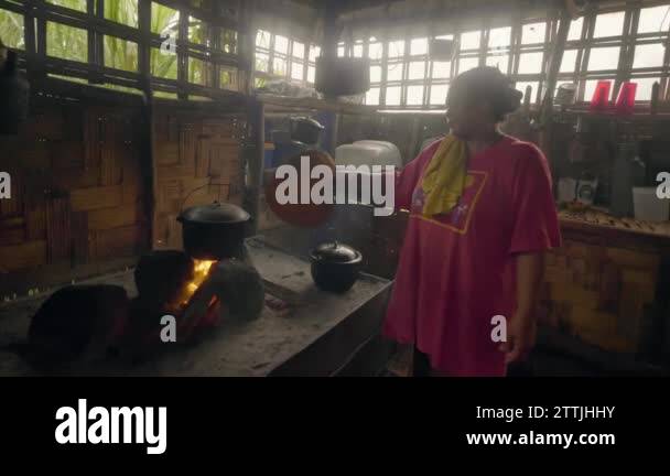Asian woman blowing fire with fan while cooking food in pot on bonfire ...