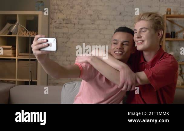 Two international gay friends, sitting on the couch, hugging, making ...