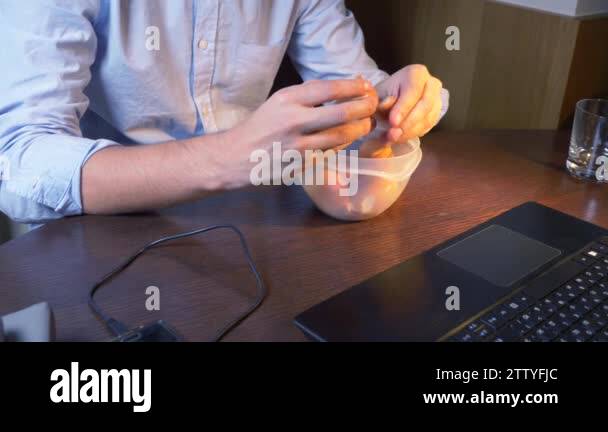 A man eats boiled chicken eggs from a food container, working on a laptop, late at night ...