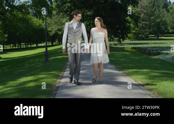 SLOW MOTION: Happy pregnant couple holding hands and walking trough green park in sunny spring ...