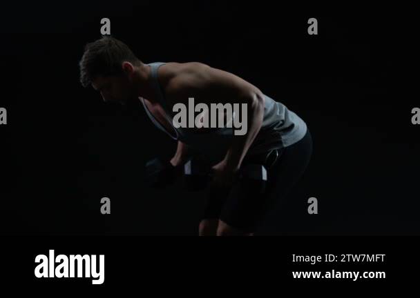 4K Fit young man with muscular physique lifting weights against black ...