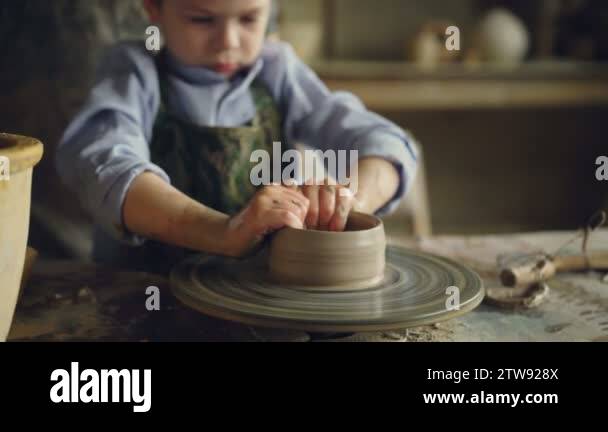 Serious boy is forming pot from clay on throwing wheel while attending ...