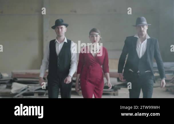 Two confident men in hats and suits and woman in red dress walking toward the camera in an ...