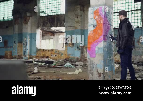 Young man graffiti artist is painting on pillar in old dirty building ...