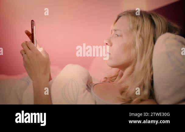Frustrated woman lying in bed typing angrily on her smartphone feeling ...