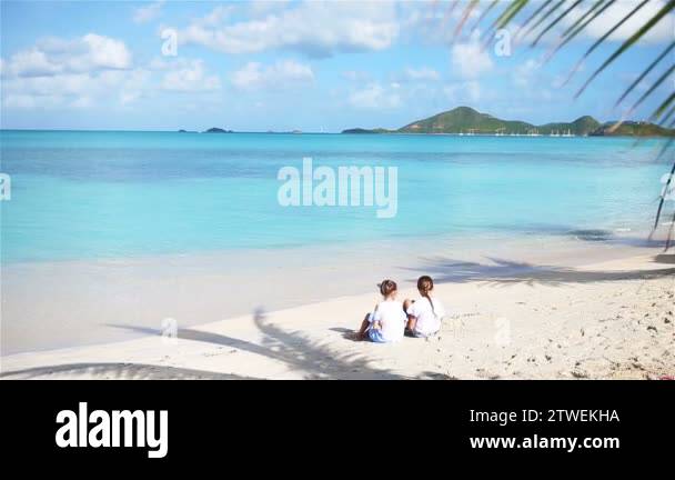 Two little girls on sandy beach. Happy kids sitting under the palm tree ...