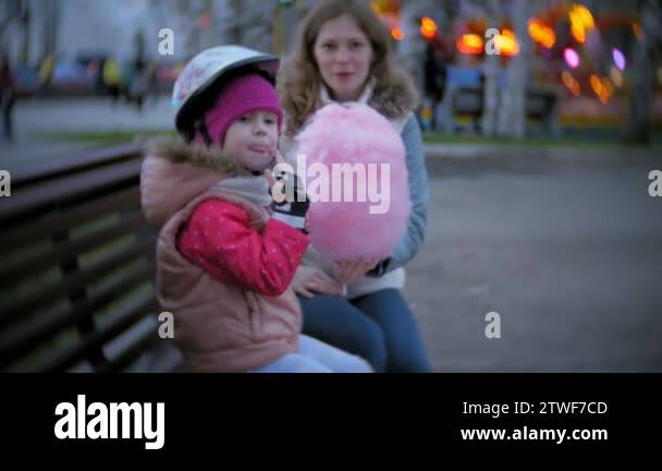 Beautiful little girl sitting on a bench in an amusement park, eating ...