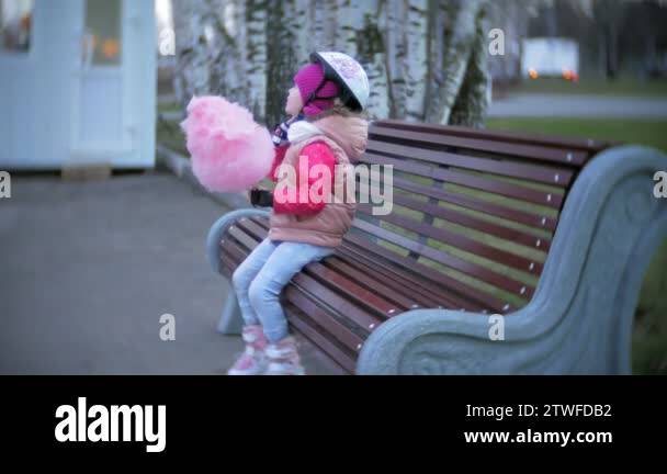 Beautiful little girl sitting on a bench in an amusement park, eating ...