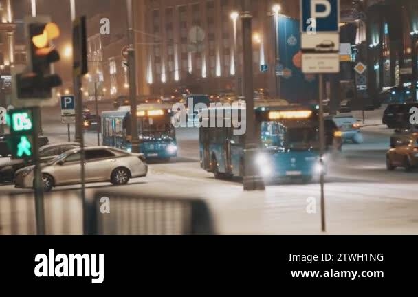 Two blue shuttle buses rides down night city street in falling snow ...