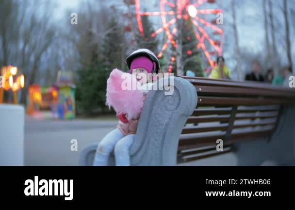 Beautiful little girl sitting on a bench in an amusement park, eating ...