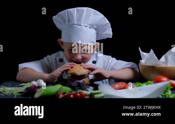 Cute boy is eating a burger while cooking. He wears chefs suit and cap ...