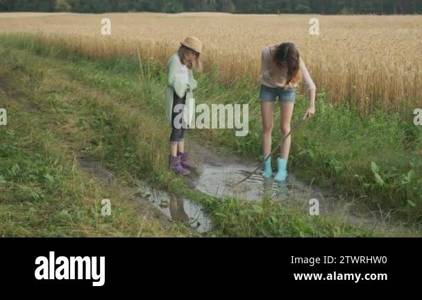 Two happy girls sisters in boots playing in puddle of rainwater on country road, summer nature ...