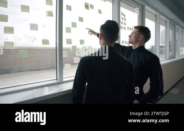 Young men are talking, pointing on notes on window in modern office ...