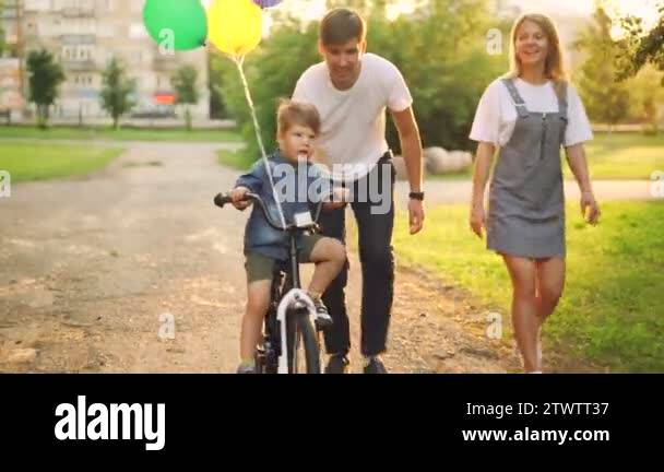 Loving parents are teaching son cute boy to ride bike in city park, child is cycling and his ...
