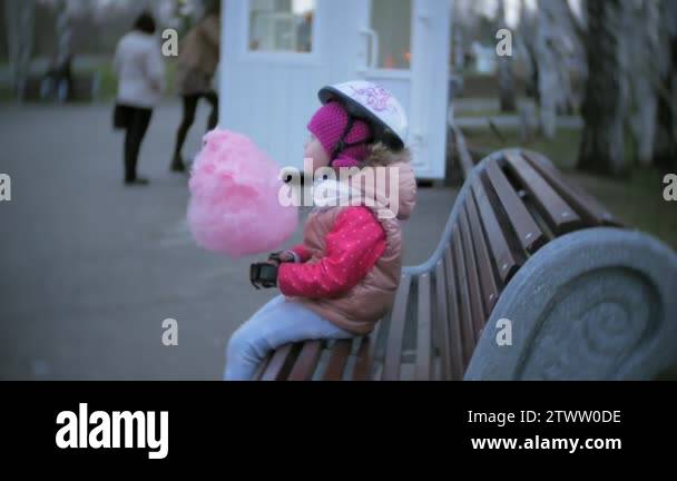 Beautiful little girl sitting on a bench in an amusement park, eating ...