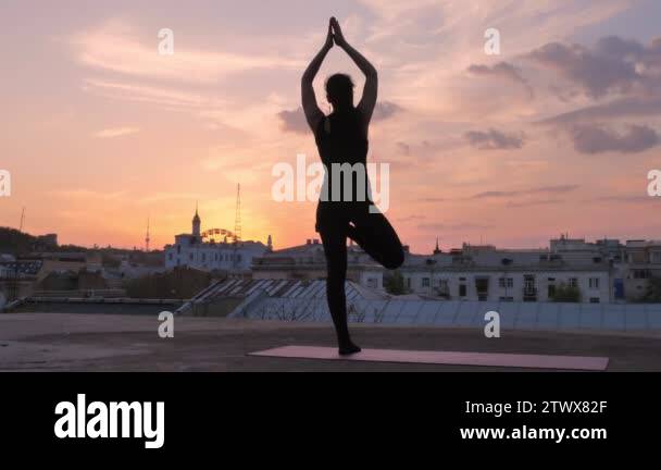 Yogi girl is standing in tree pose on rooftop in summer on sunset ...