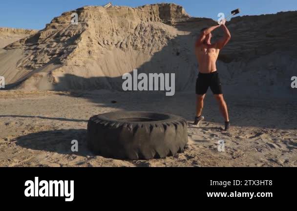 Muscle athlete strongman man hits a hammer on a huge wheel in the sandy ...