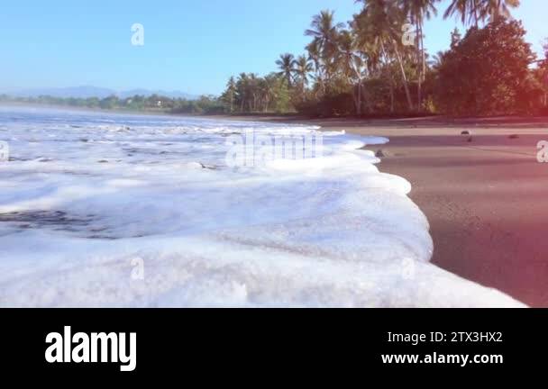 SLOW MOTION CLOSE UP LENS FLARE: Foamy ocean waves washing black sandy ...