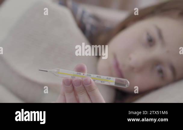 Teenage girl wrapped in a blanket lying in bed showing thermometer to the camera, fever, video ...