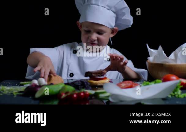 Cute boy is eating a burger while cooking. He wears chefs suit and cap ...