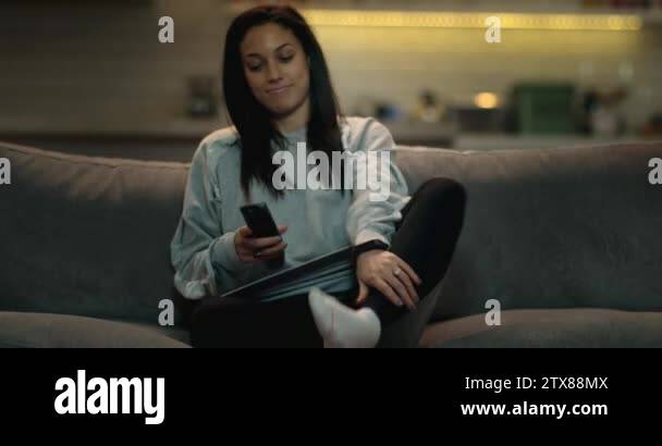 Woman sits down on a sofa, crosses her legs and turns the tv on using a ...