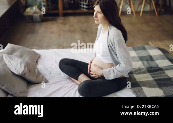 Pregnant lady is caressing her tummy sitting on double bed at home and ...