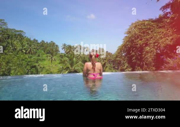 AERIAL CLOSE UP LENS FLARE: Flying above young woman in pink bikini hanging in infinity pool and ...