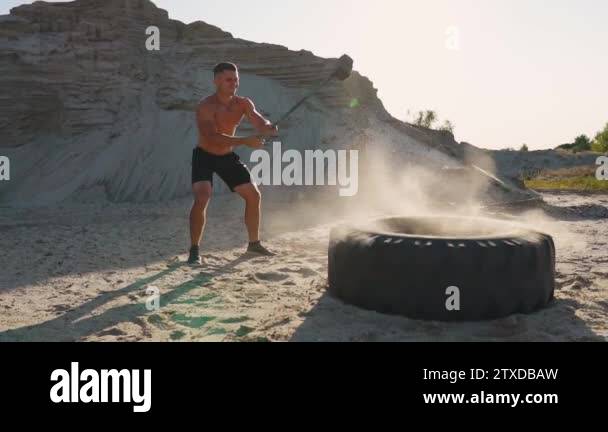 Muscle athlete strongman man hits a hammer on a huge wheel in the sandy ...