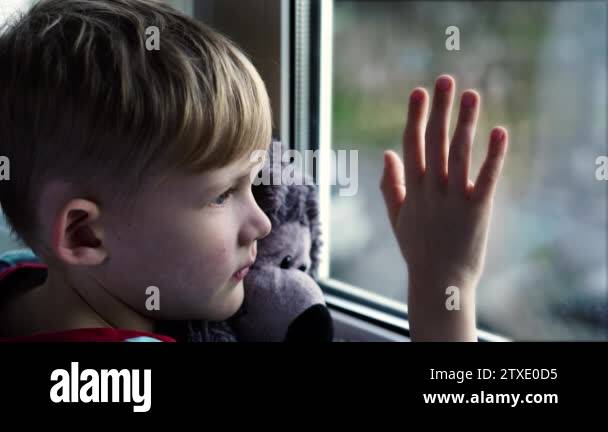 Little boy stares sadly out of a window with a teddy bear in her arms ...