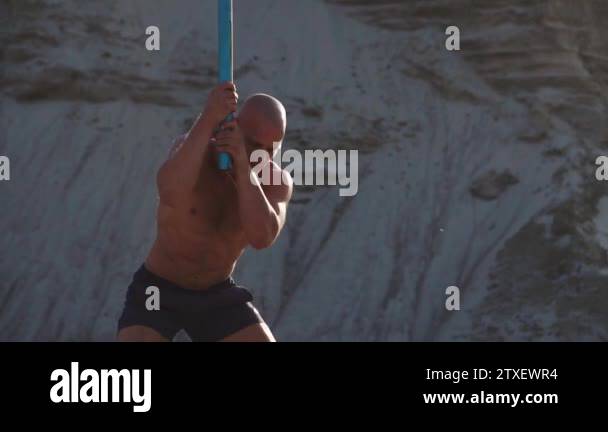 Bald man strongman hits a hammer on a huge wheel in the sandy mountains ...