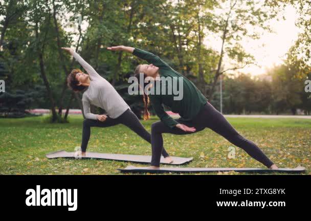 Two pretty women are doing yoga outdoors in park on mats practising asanas and breathing fresh ...