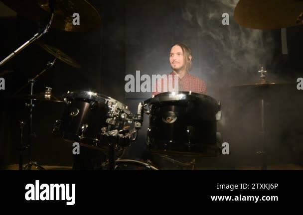 A long-haired drummer sitting behind a drum kit is hovering with a vape ...
