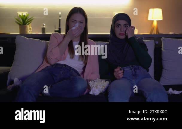 Two girls watching sad film on couch, crying and wiping tears, leisure ...