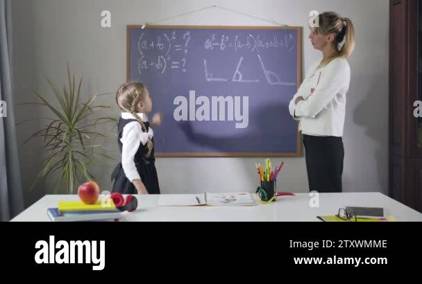 Pretty Caucasian girl in elegant school uniform and middle aged tutor standing at blackboard and ...