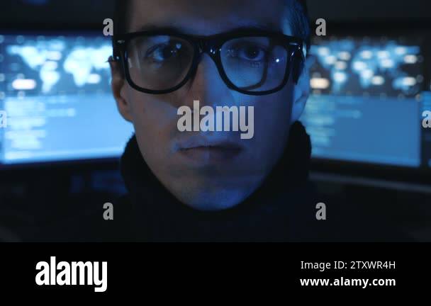 Portrait of Hacker programmer in glasses looks at camera while blue code characters reflect on ...