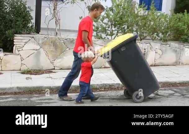 Father and son together push big trash can on wheels on the street ...