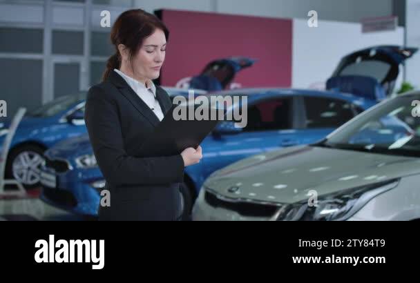 Portrait of Caucasian female car dealer standing in dealership with document holder and thinking ...