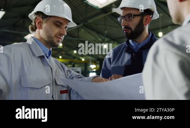 Male engineers in helmets with big paper drawings talking, discussing ...