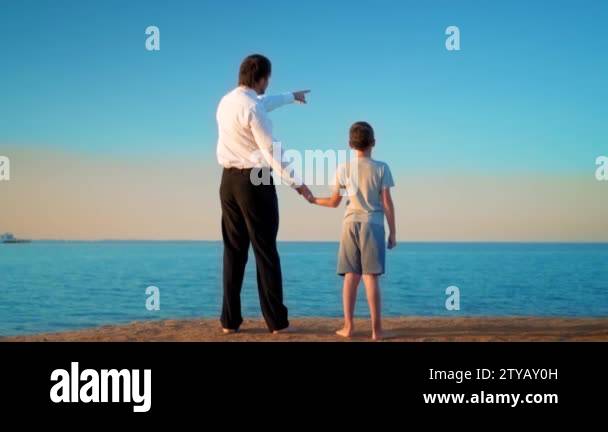 The father and son stand, by the hand, on the seashore, father points ...