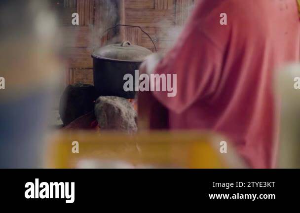 Woman blowing fire with fan while cooking food on bonfire in pot in asian village. Woman cooking ...